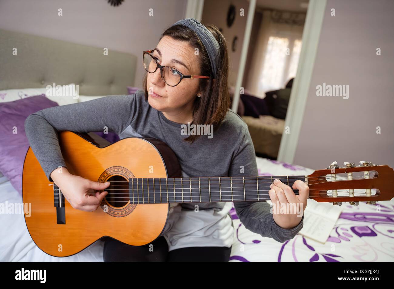 Woman strums an acoustic guitar on a cozy bed, lost in thought ...