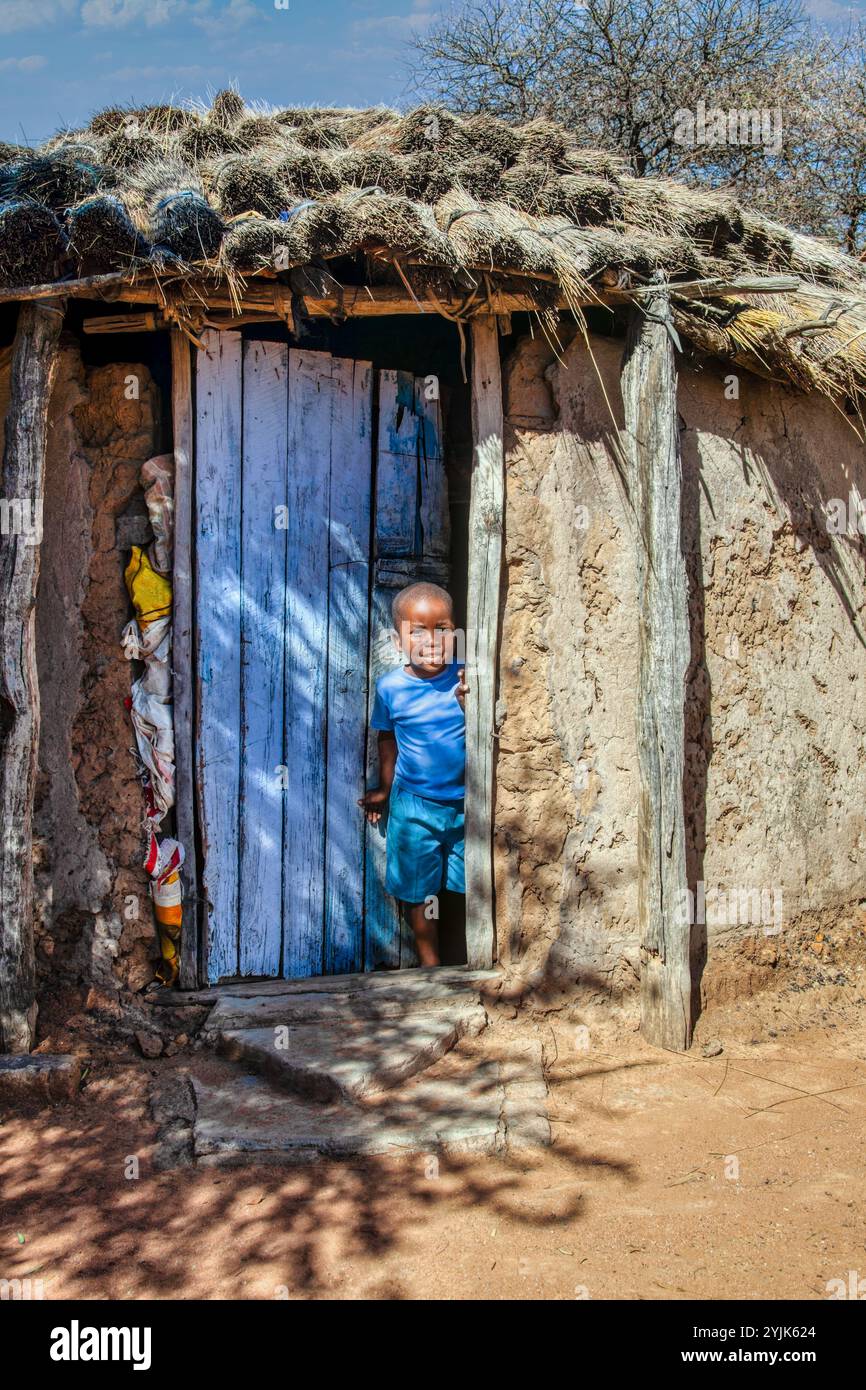 Single village african boy child standing in front of mud hut with thatched roof and wooden door ...