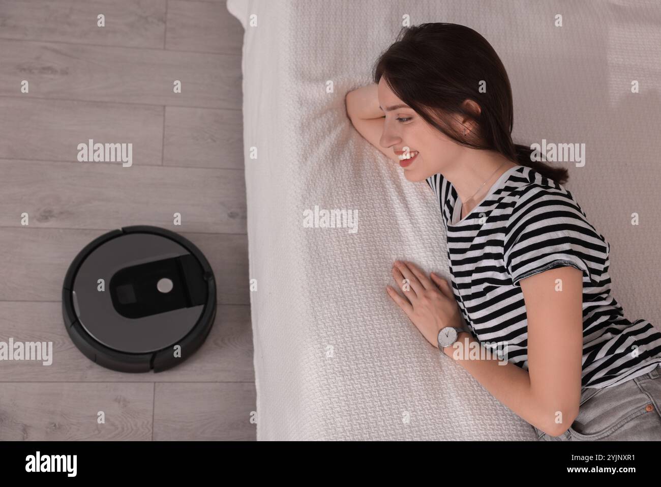 Smiling young woman resting on bed while robotic vacuum cleaner ...