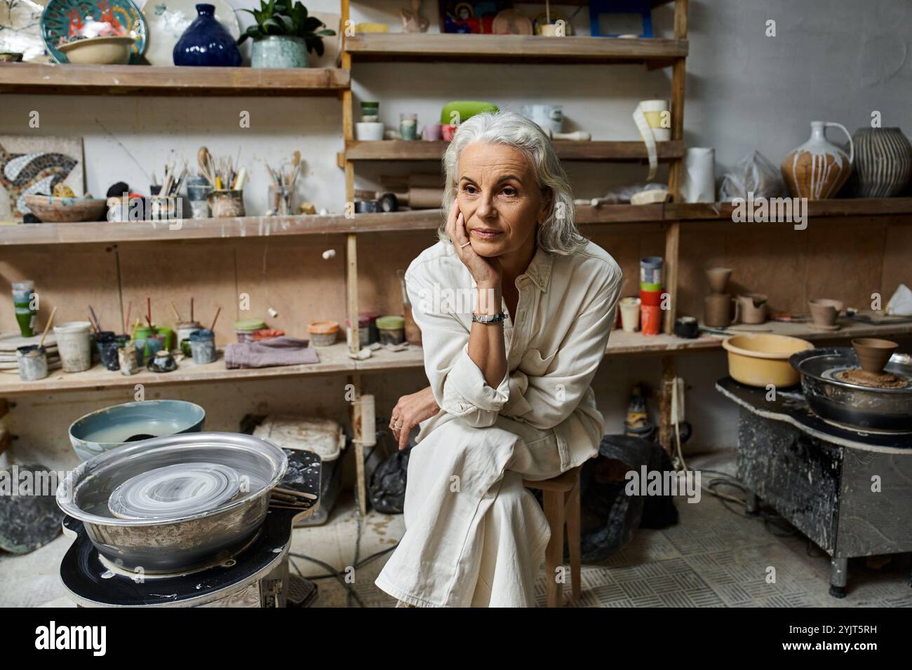 The mature woman sits thoughtfully in her pottery studio, surrounded by ...