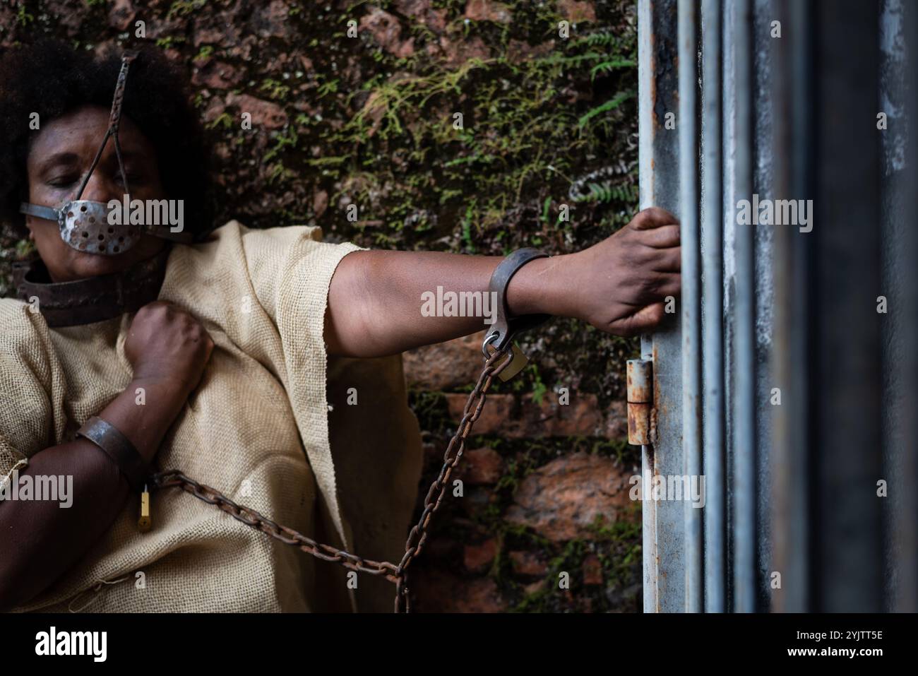 Black woman dressed as a slave, chained with an iron mask on her mouth and neck against a brick ...