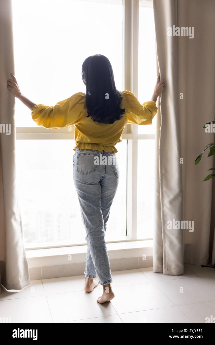 Back view of woman parting curtains at large window Stock Photo - Alamy