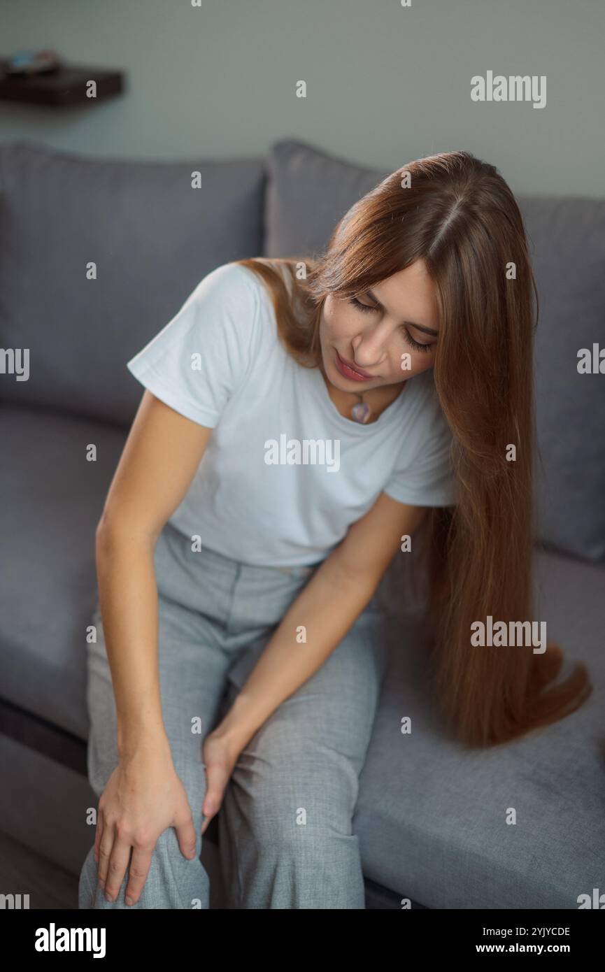 Young woman sitting on sofa experiencing discomfort in her knee while ...