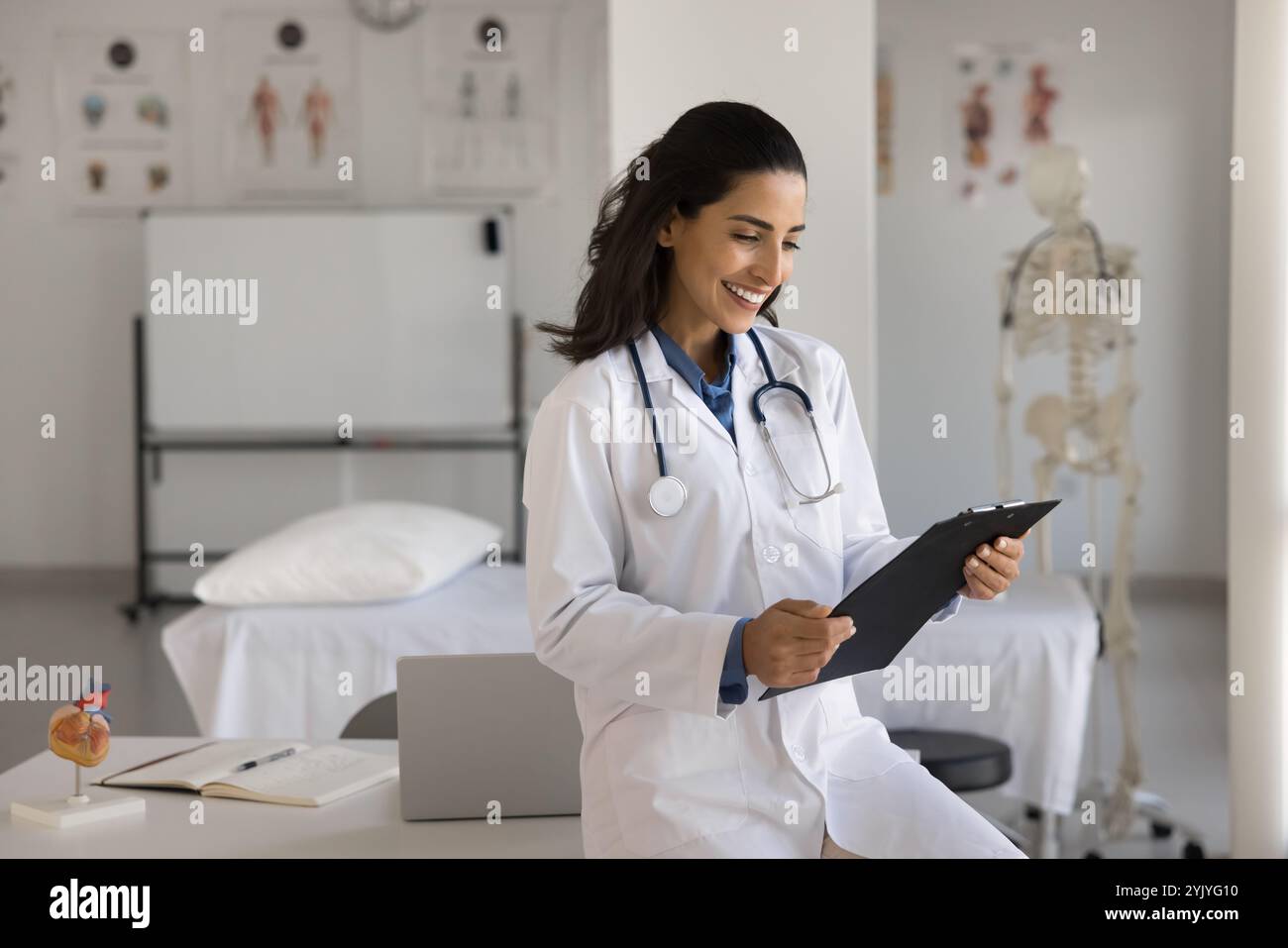 Smiling Brazilian female cardiologist holding clipboard standing in ...