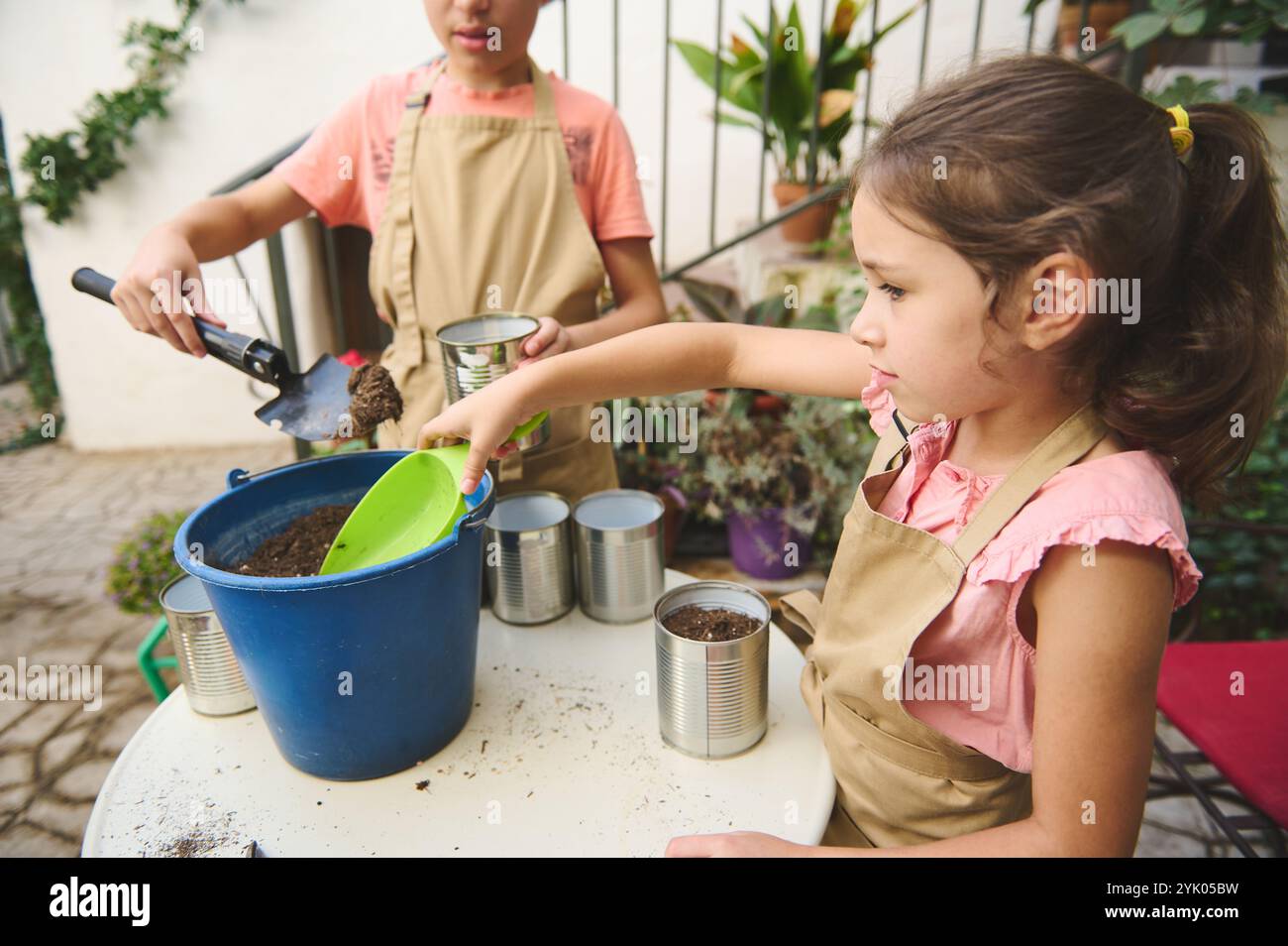 Two children wearing aprons are planting seeds in metal containers on a ...