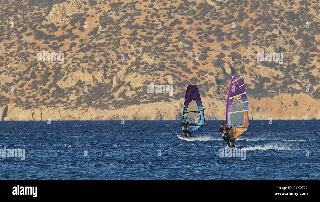 Two windsurfers in front of a rocky coast and a clear blue sky ...