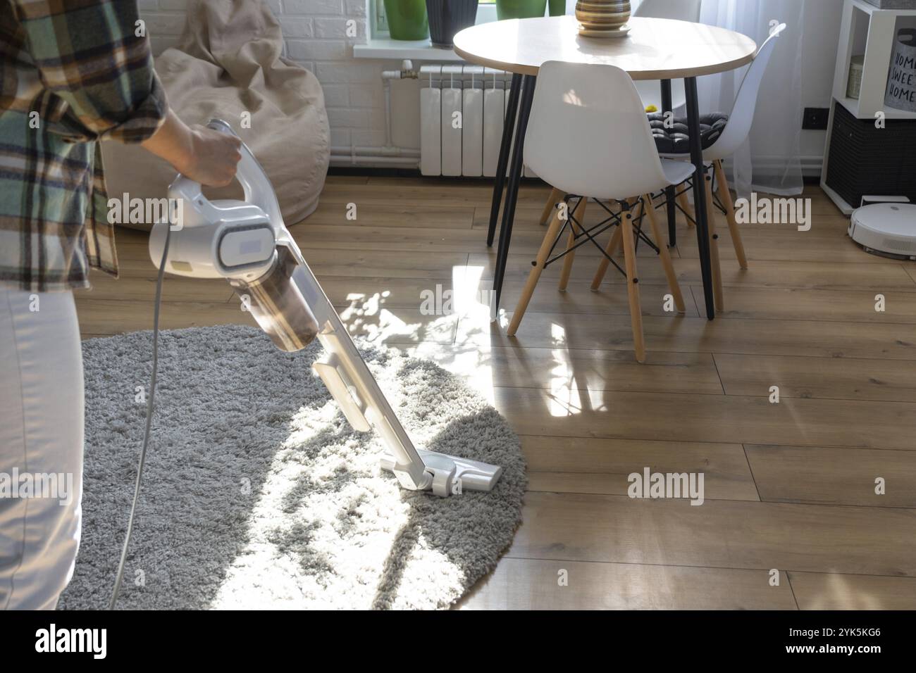 A woman vacuums a round carpet in a house among house plants with a ...