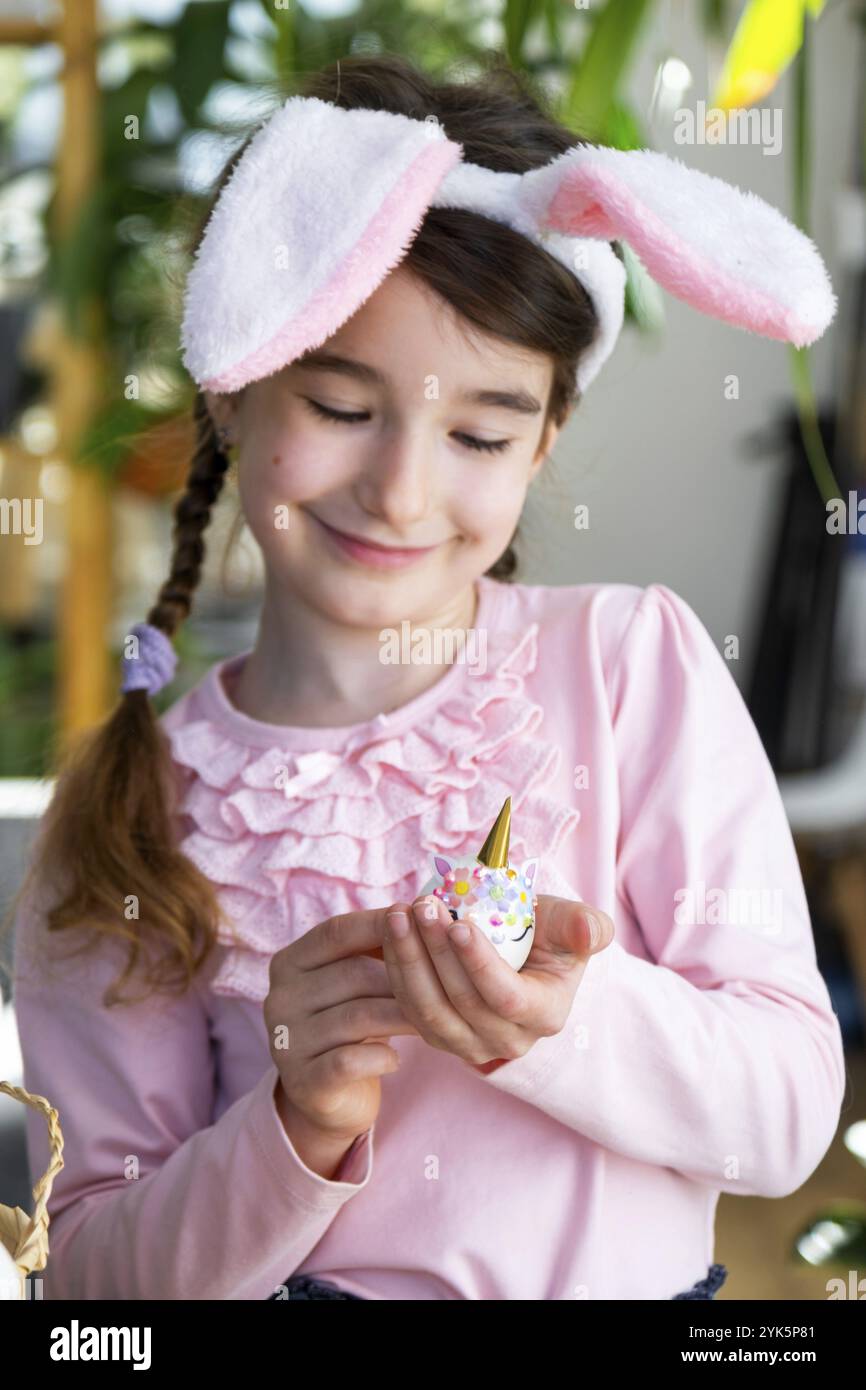 A cute girl with pink bunny ears makes an Easter craft, decorates an ...
