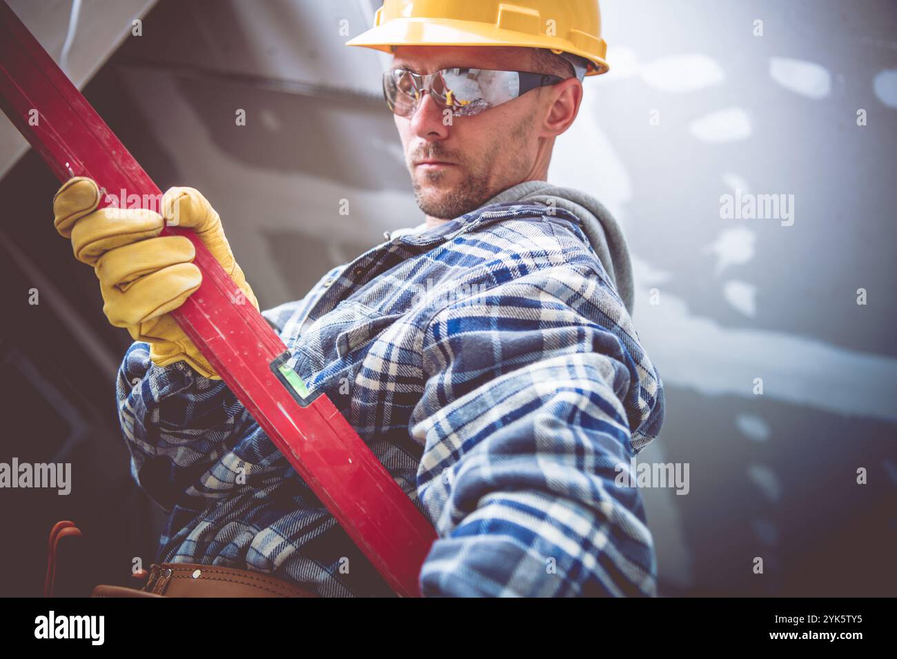 A construction worker in a plaid shirt and hard hat is measuring a wall ...