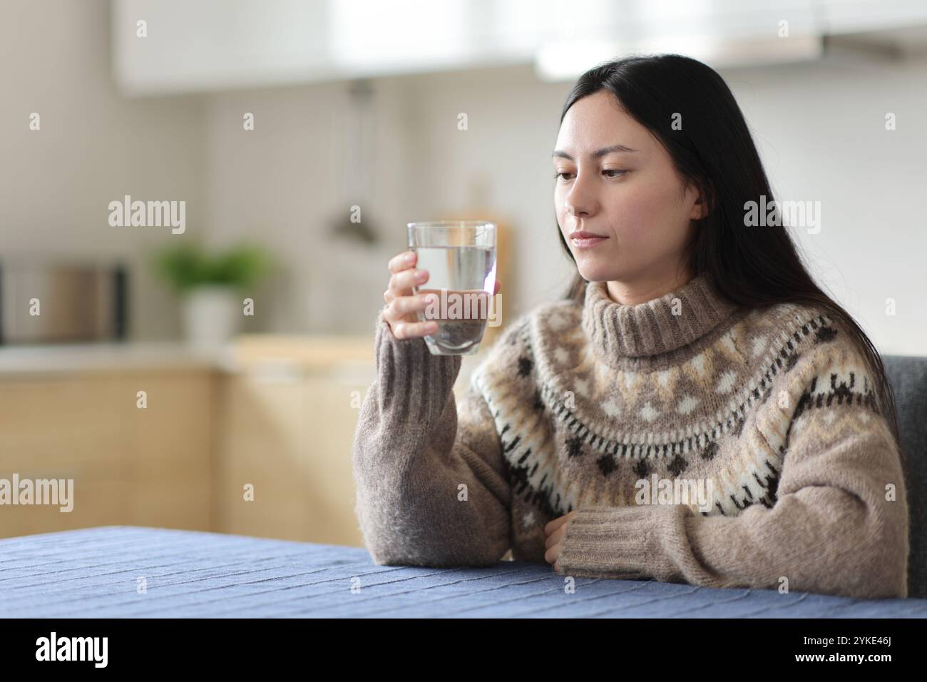 Serious asian woman in winter drinking water from glass in the kitchen ...