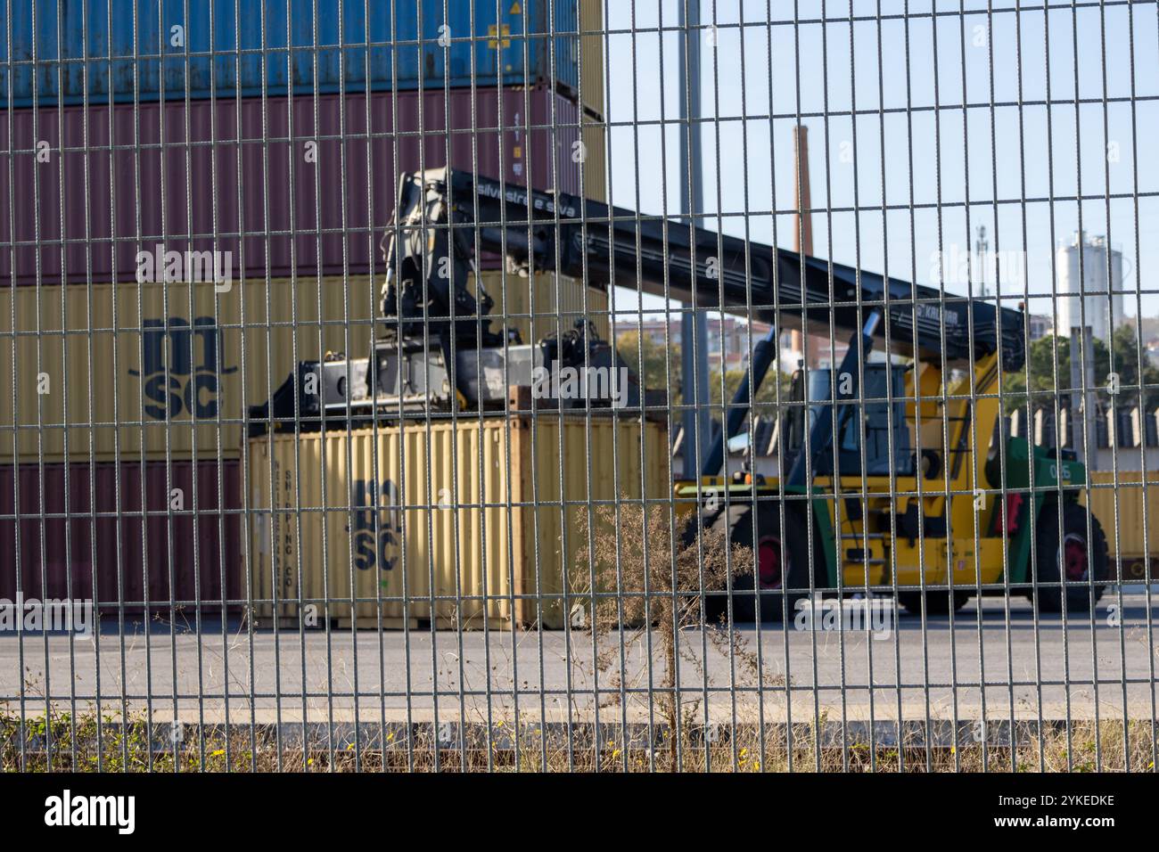 Reach stacker lifting a shipping container behind a wire mesh fence in ...