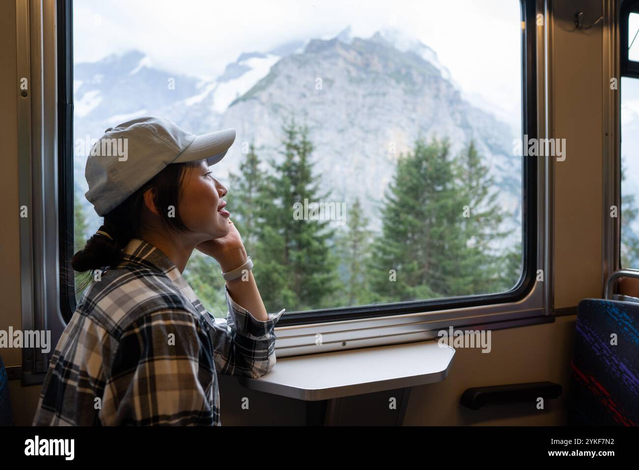 A young asian woman wearing a cap gazes out of a train window, admiring the majestic mountain ...