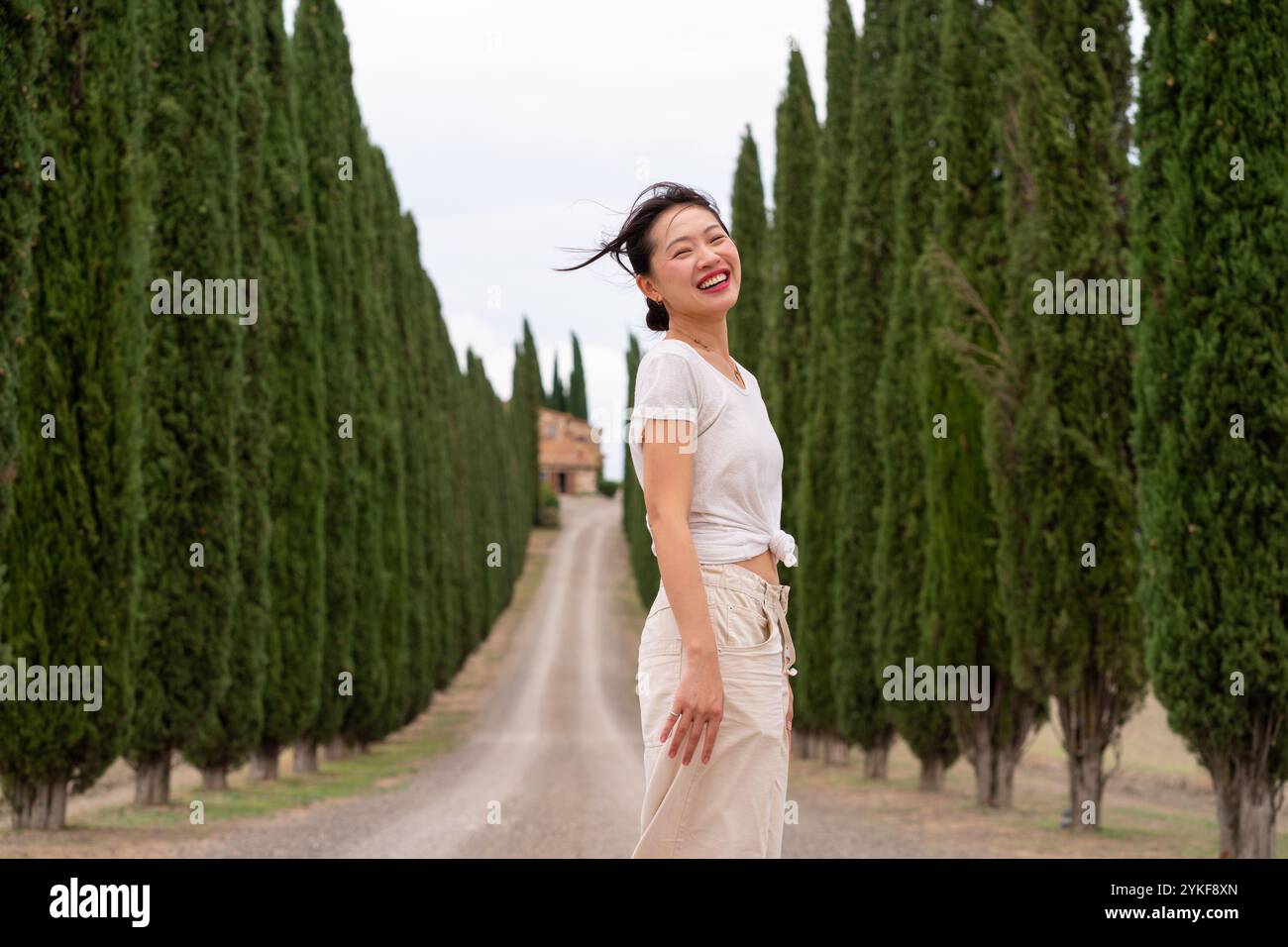 A joyful Asian woman laughs brightly as she stands on a gravel path lined with tall cypress ...