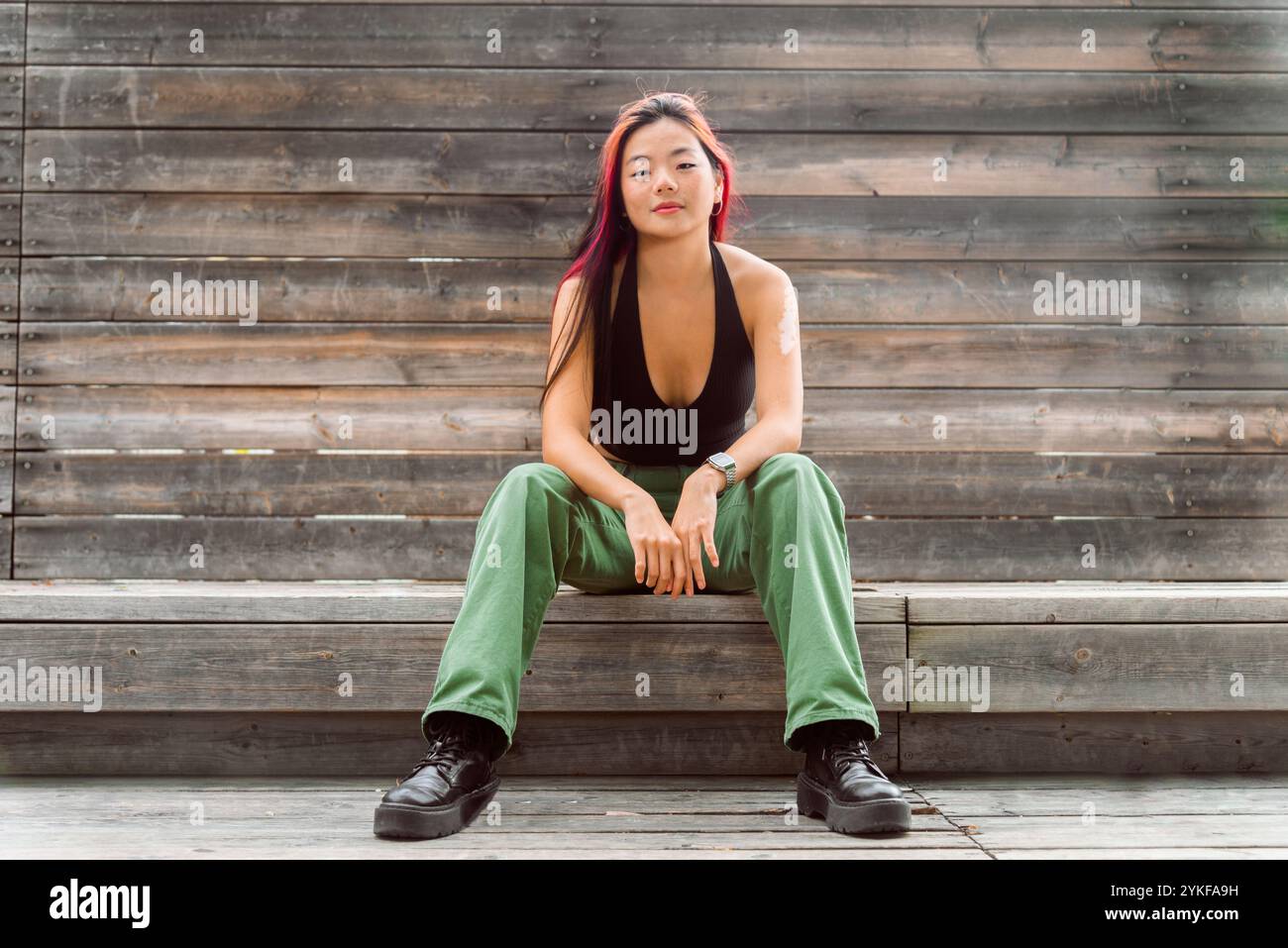 Asian woman sitting casually on wooden steps, wearing green pants and a ...