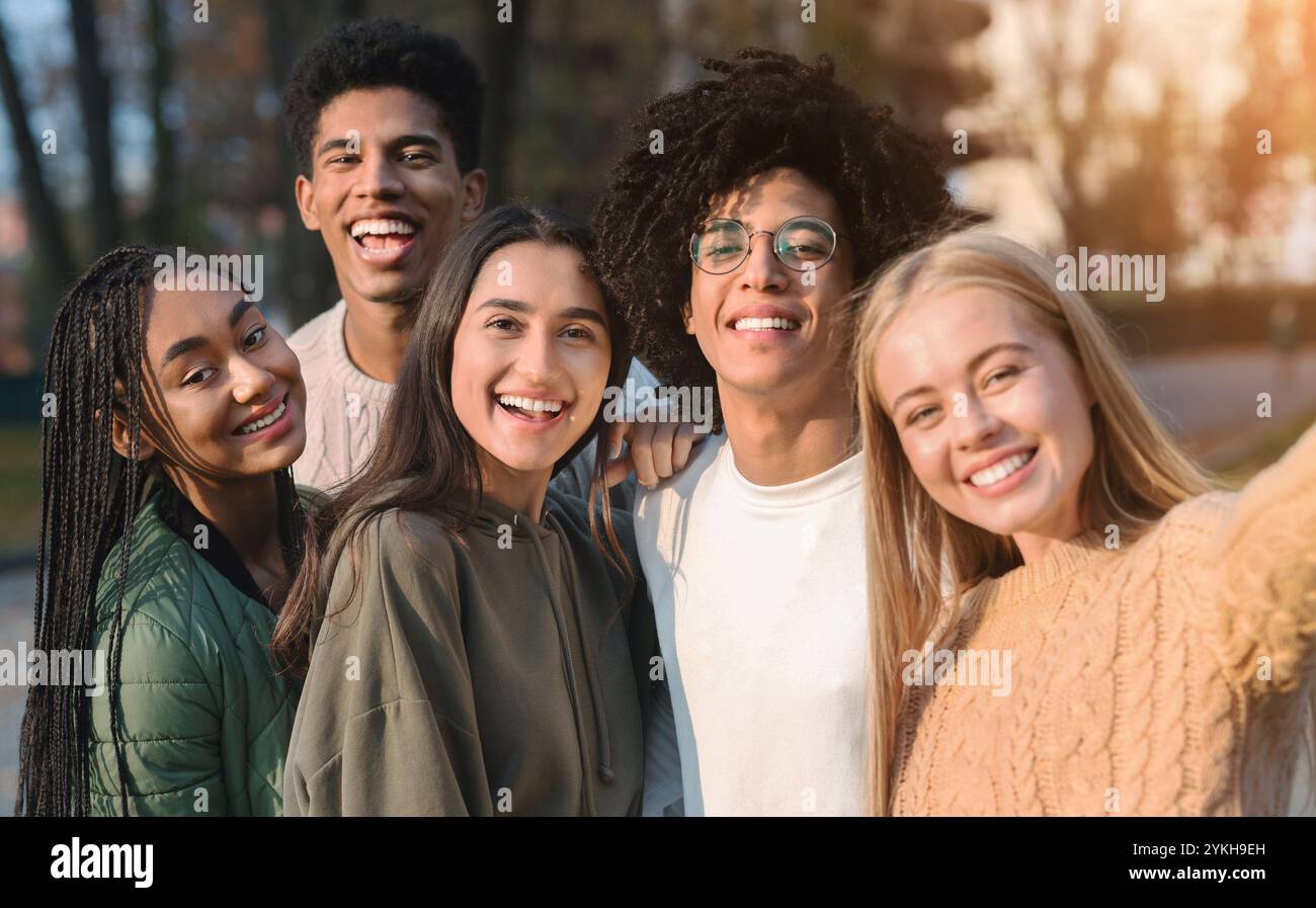 Positive multiracial teen friends taking selfie while walking in park ...