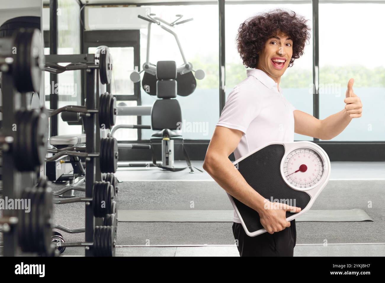 Young man holding a weight scale at a gym and gesturing a thumb up sign ...