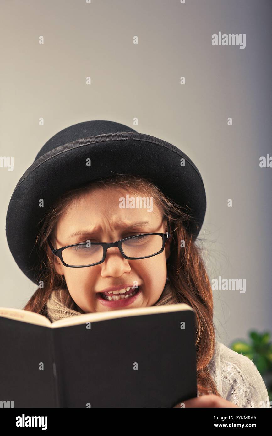 Little girl wearing a bowler hat and glasses is reading a book and ...