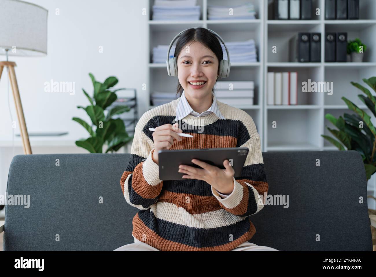 A cheerful young woman enjoys using her tablet in a stylish home office, surrounded by plants ...