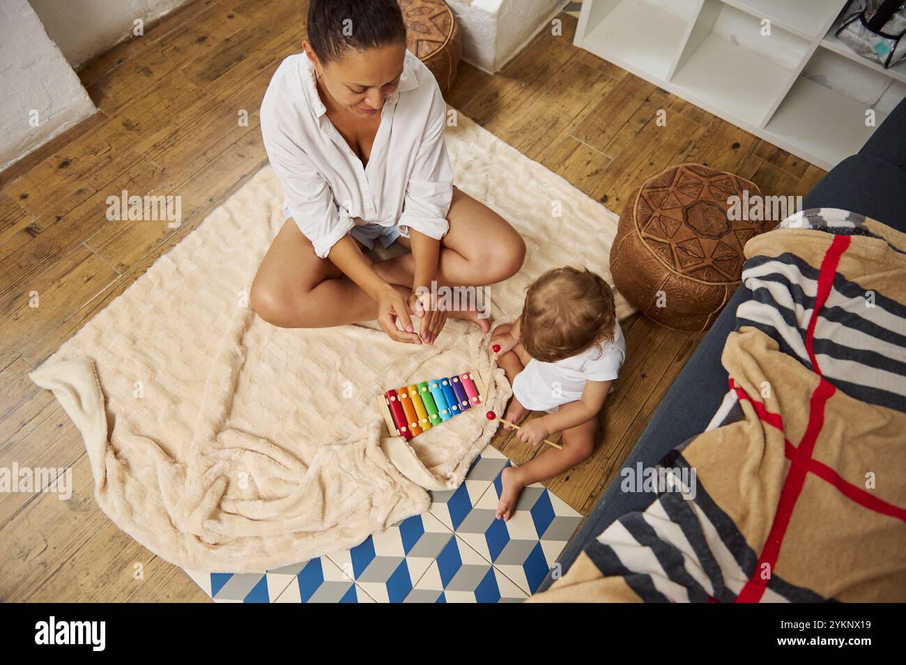 A joyful mother and child play together at home, seated on a cozy rug. The young child engages ...