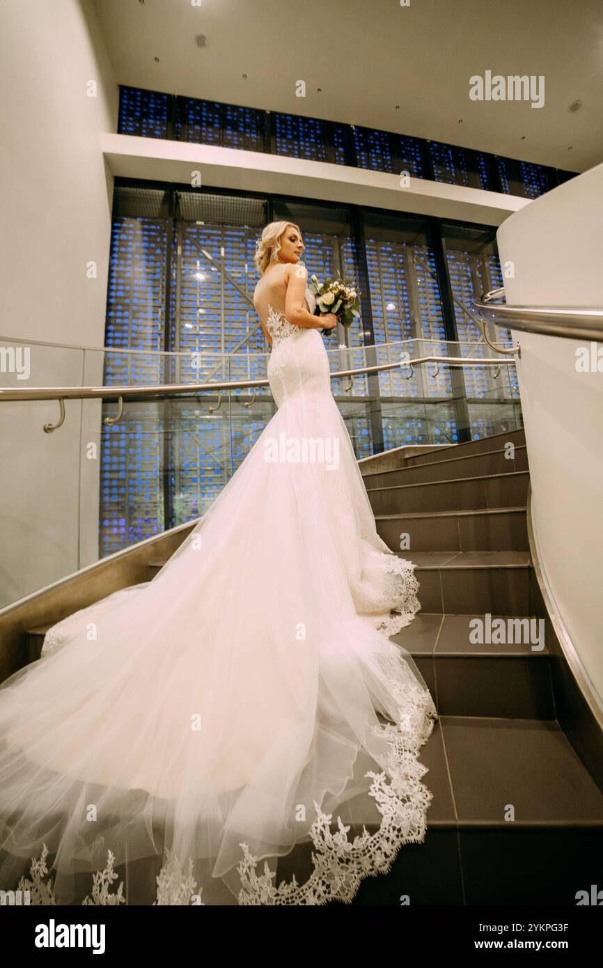 A stunningly beautiful bride gracefully poses on a magnificent staircase, showcasing her ...