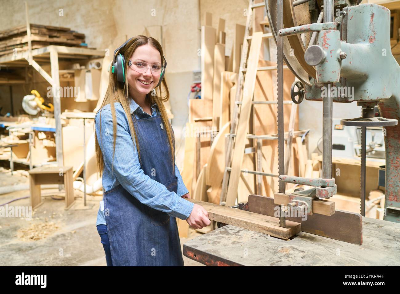 A young woman wearing safety goggles and headphones operates a bandsaw ...