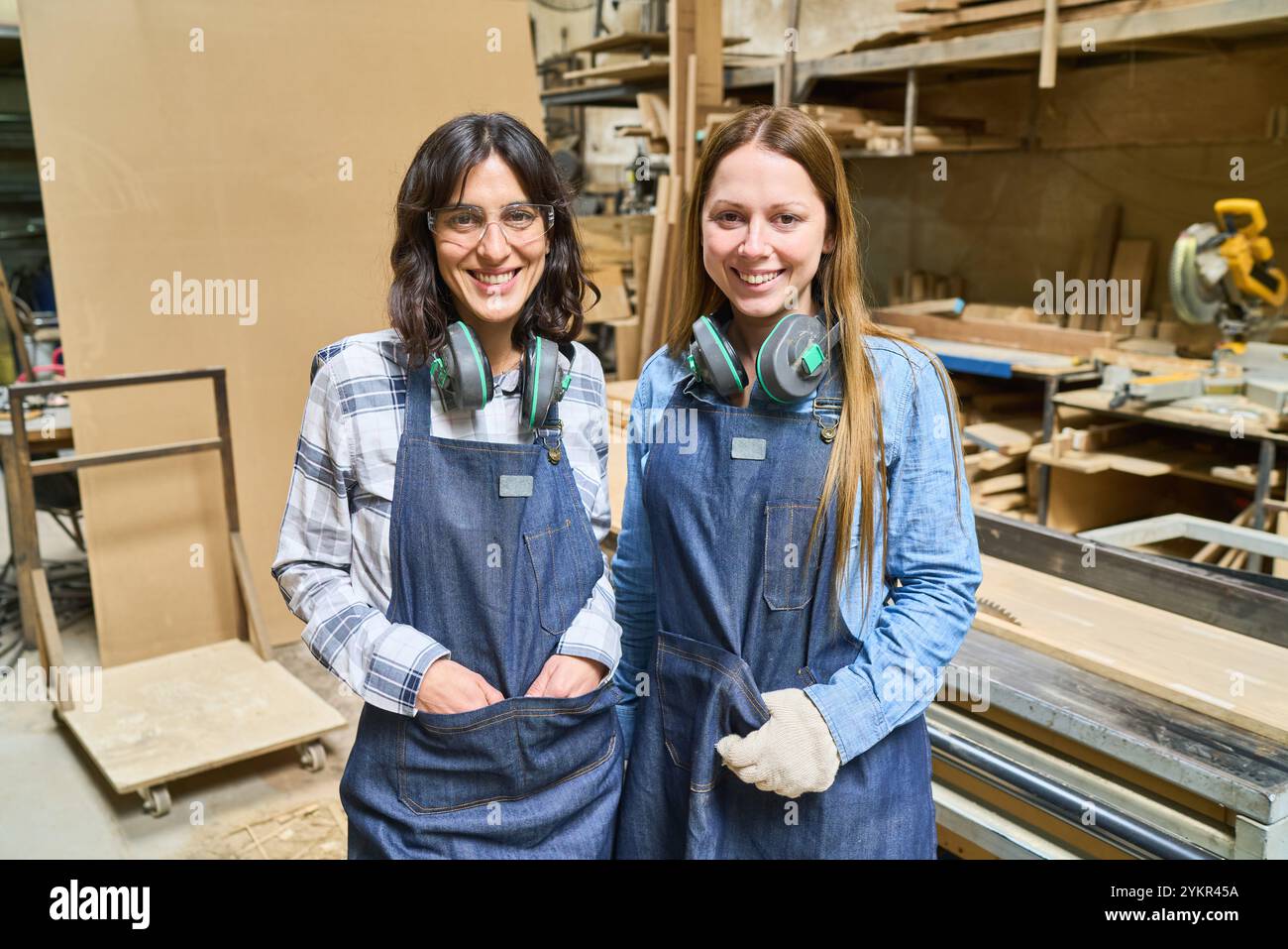 Two young women in a lumberyard, wearing denim aprons and safety gear ...
