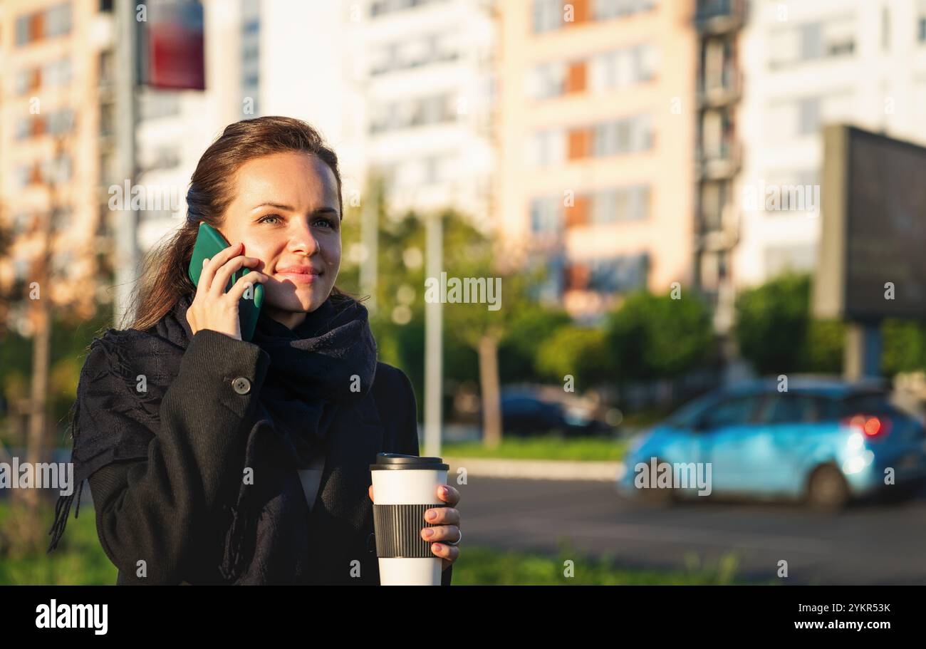 Woman walking on city street holding a coffee mug and talking on her ...