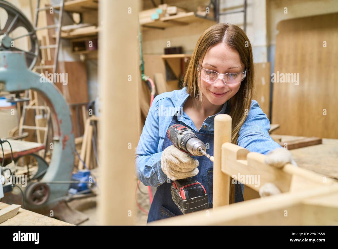 A young woman wearing safety gear uses a drill in a lumberyard workshop, embodying the spirit of ...