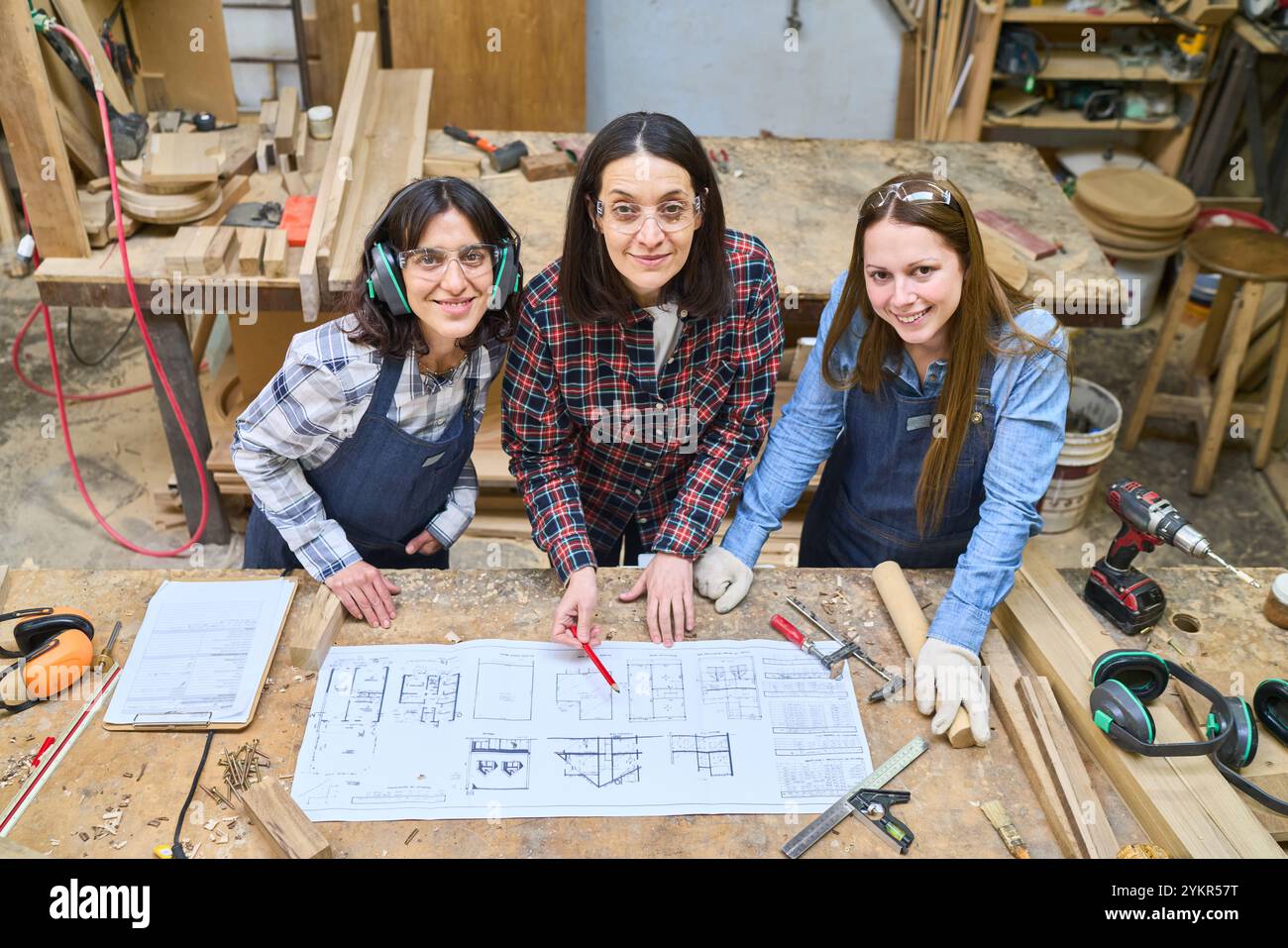 Three women collaborate on a woodworking project, examining blueprints ...