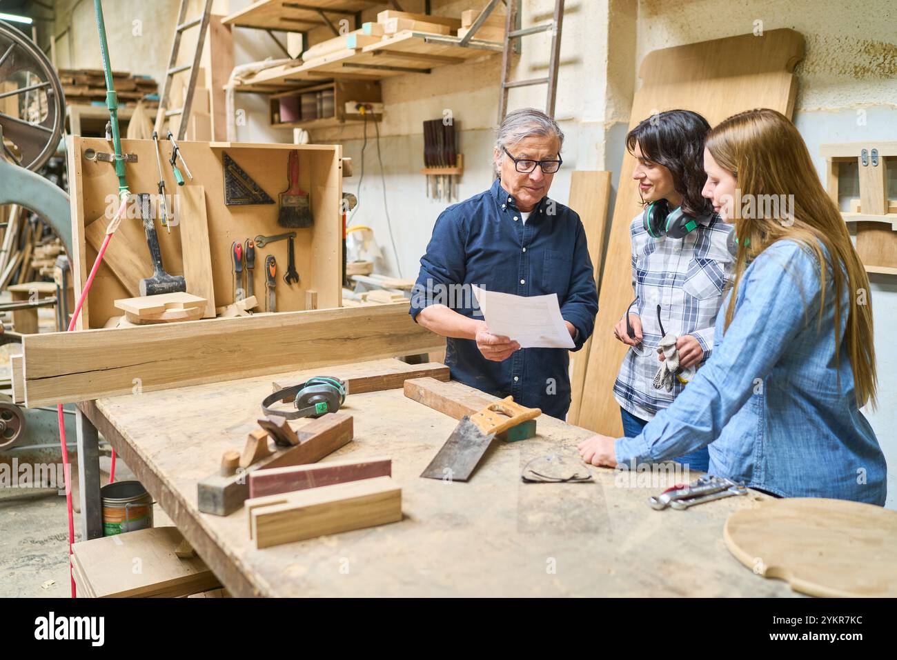 An experienced worker guides two younger colleagues in a lumberyard, showcasing teamwork and ...