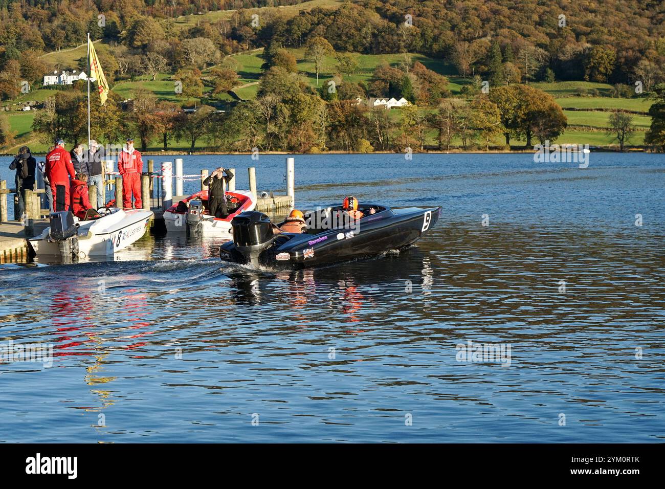 Group of people watching 2 others in a black power boat leaving a jetty or dock on Coniston ...