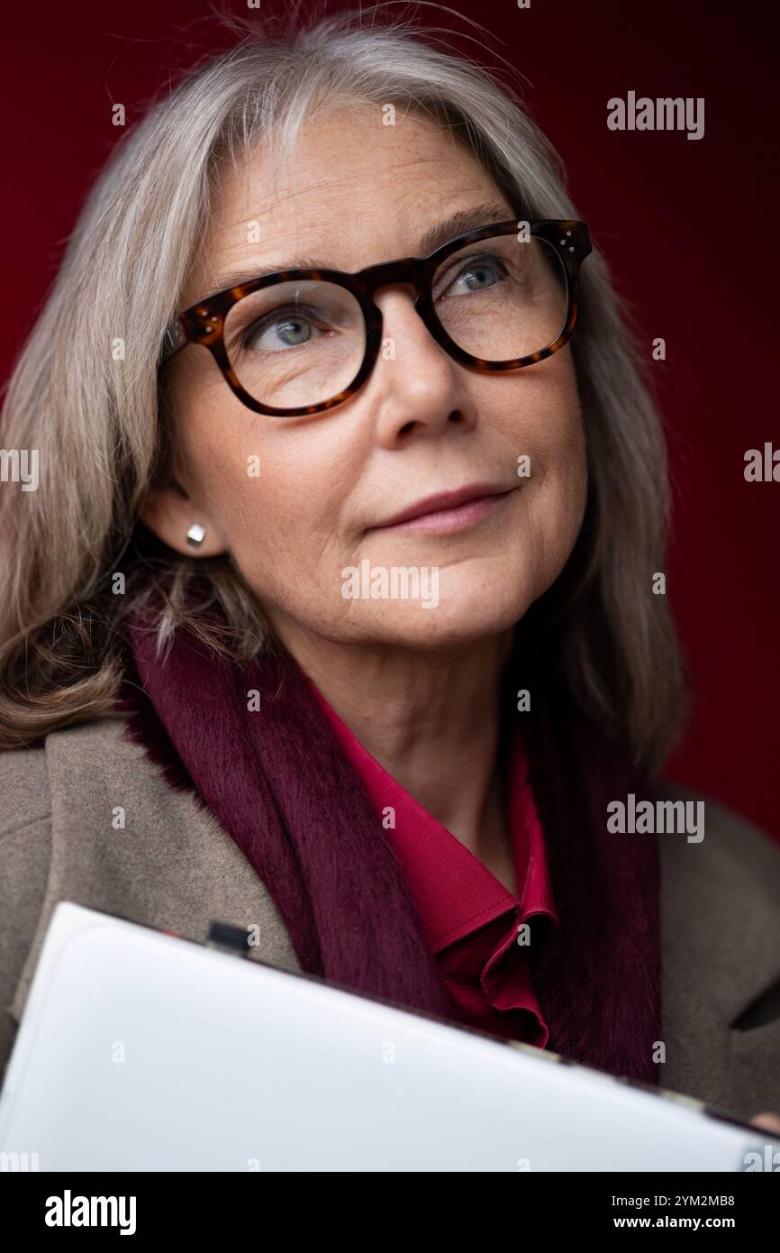 An older woman with glasses reflects thoughtfully, holding a notepad ...