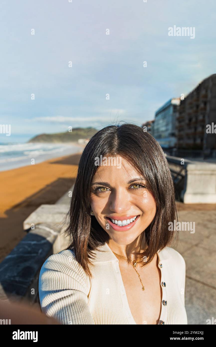Woman with a bright smile takes a selfie along a beach promenade ...