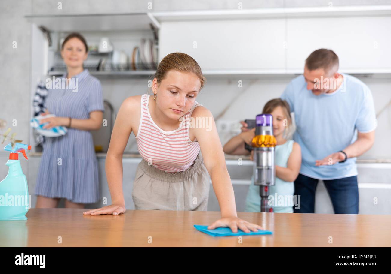 Bored teenage girl wiping table while cleaning apartment with family ...
