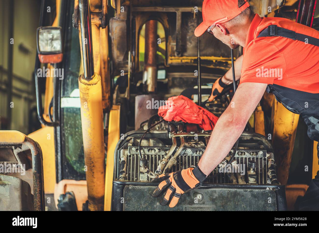 A mechanic in bright orange attire works on the engine of yellow construction equipment inside a ...