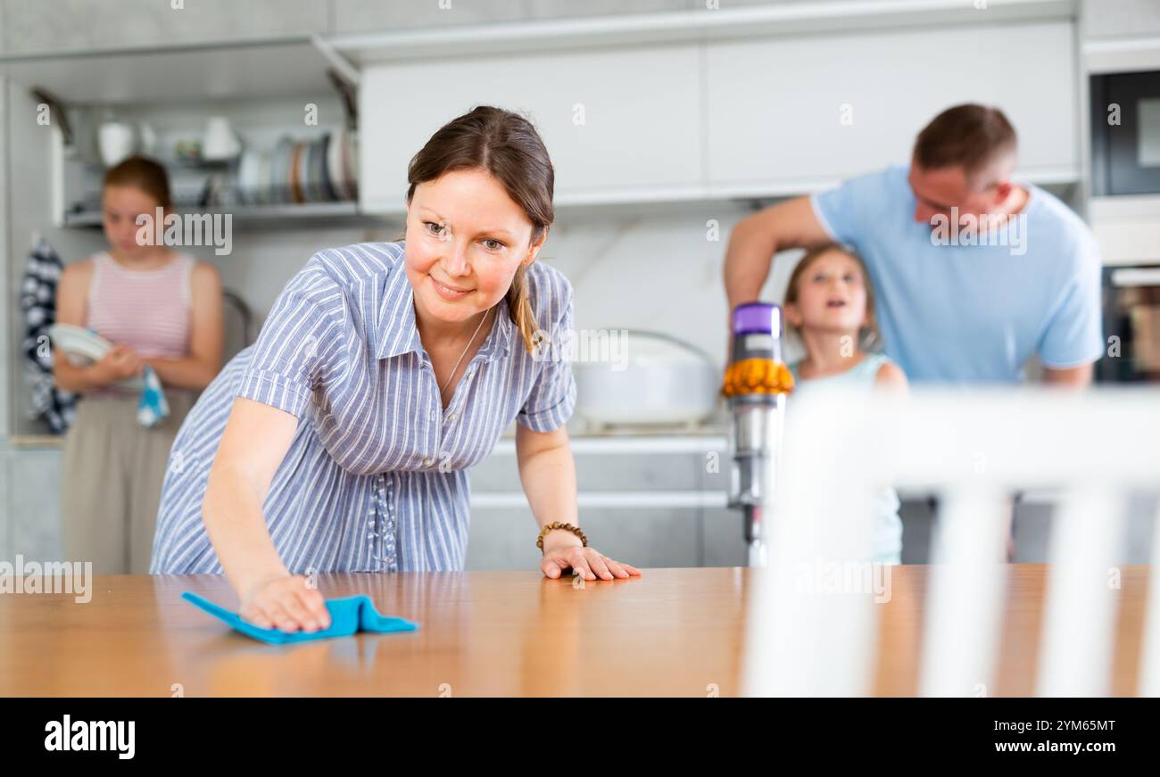 Woman rubbing table in kitchen while cleaning house with family Stock ...