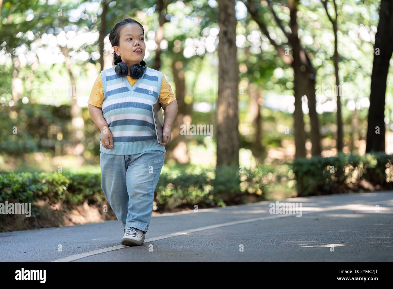 Confident Asian woman with dwarfism walking in park with headphones ...