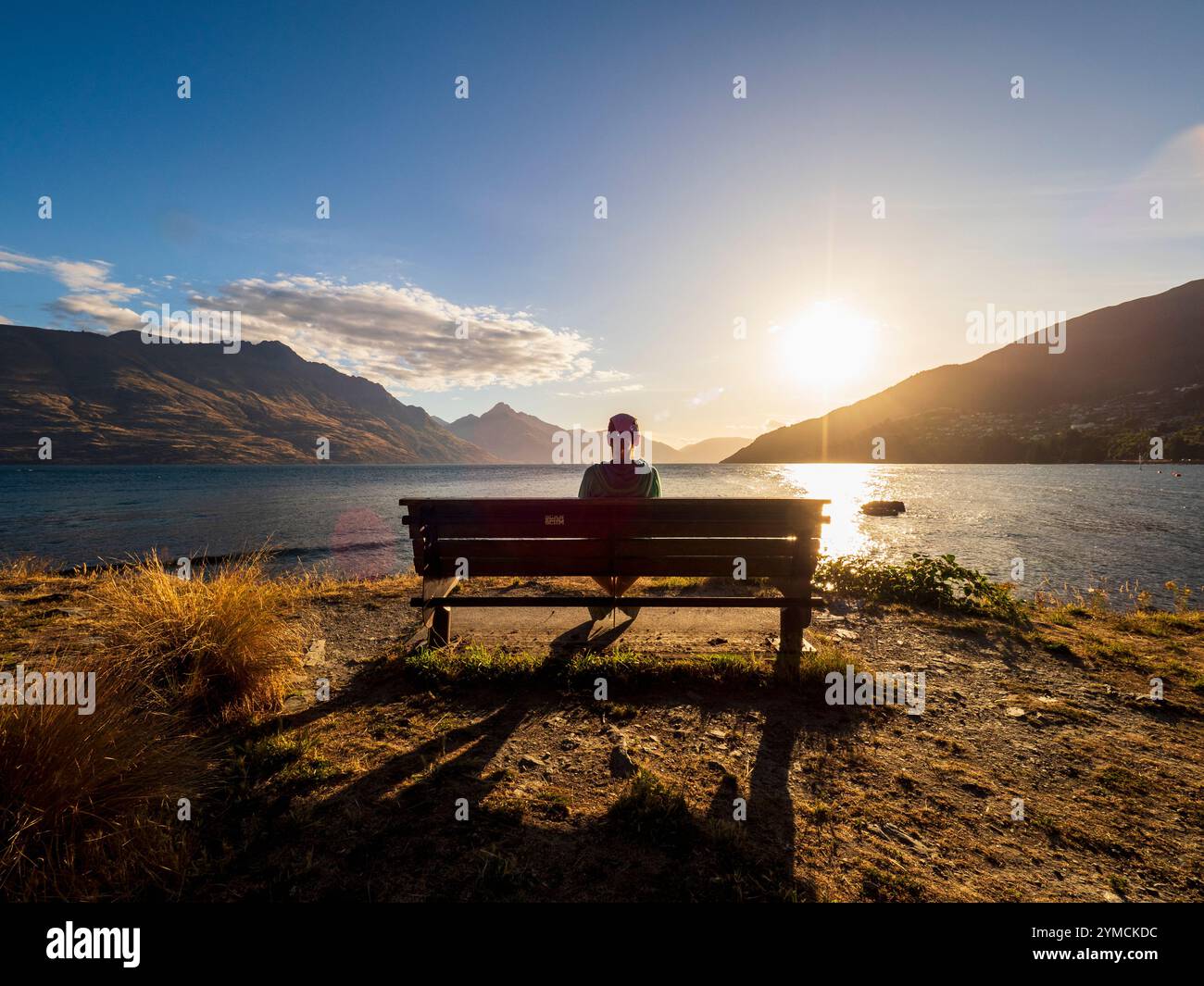 Rear view of woman sitting on bench, facing Lake Wakatipu at sunset ...