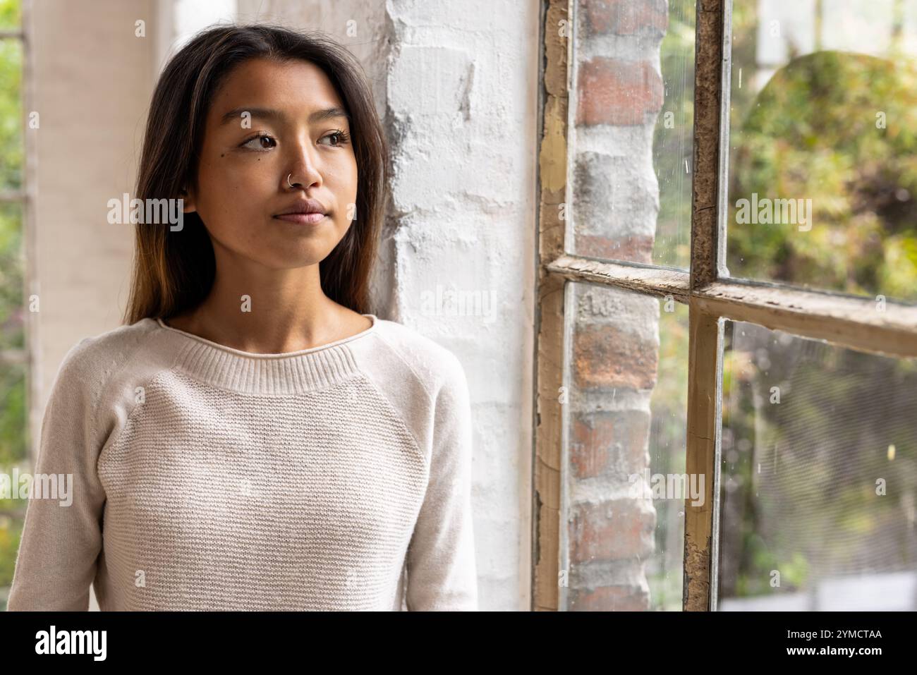 Asian female teenager standing by window, looking outside thoughtfully ...
