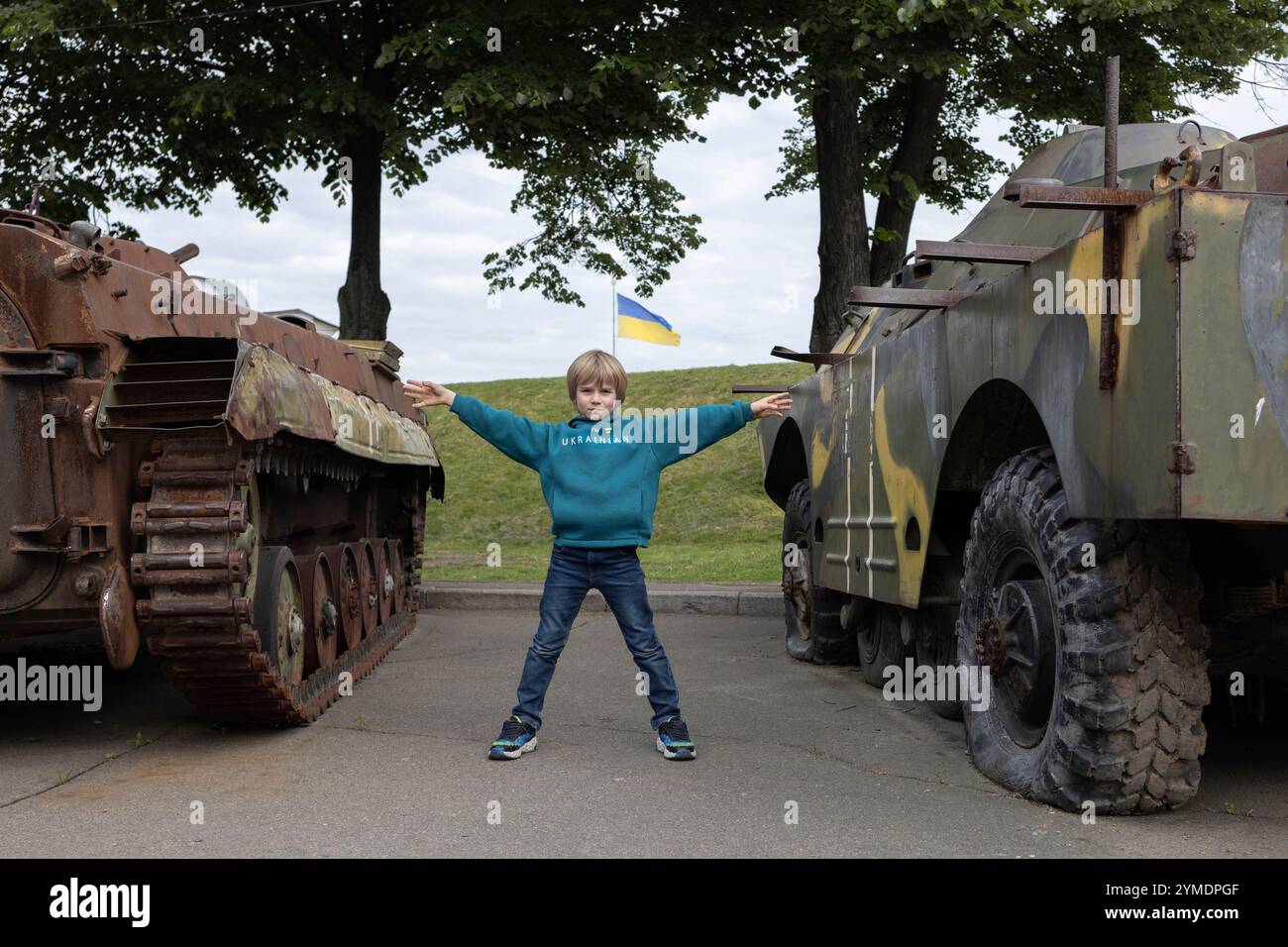boy stands between two military armored vehicles, tanks, behind him is ...