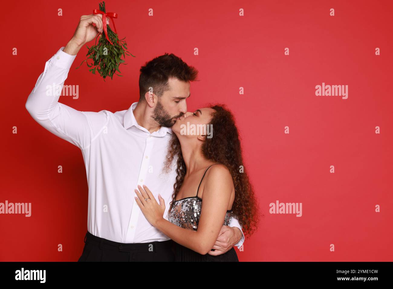 Happy couple kissing under mistletoe bunch on red background. Space for ...