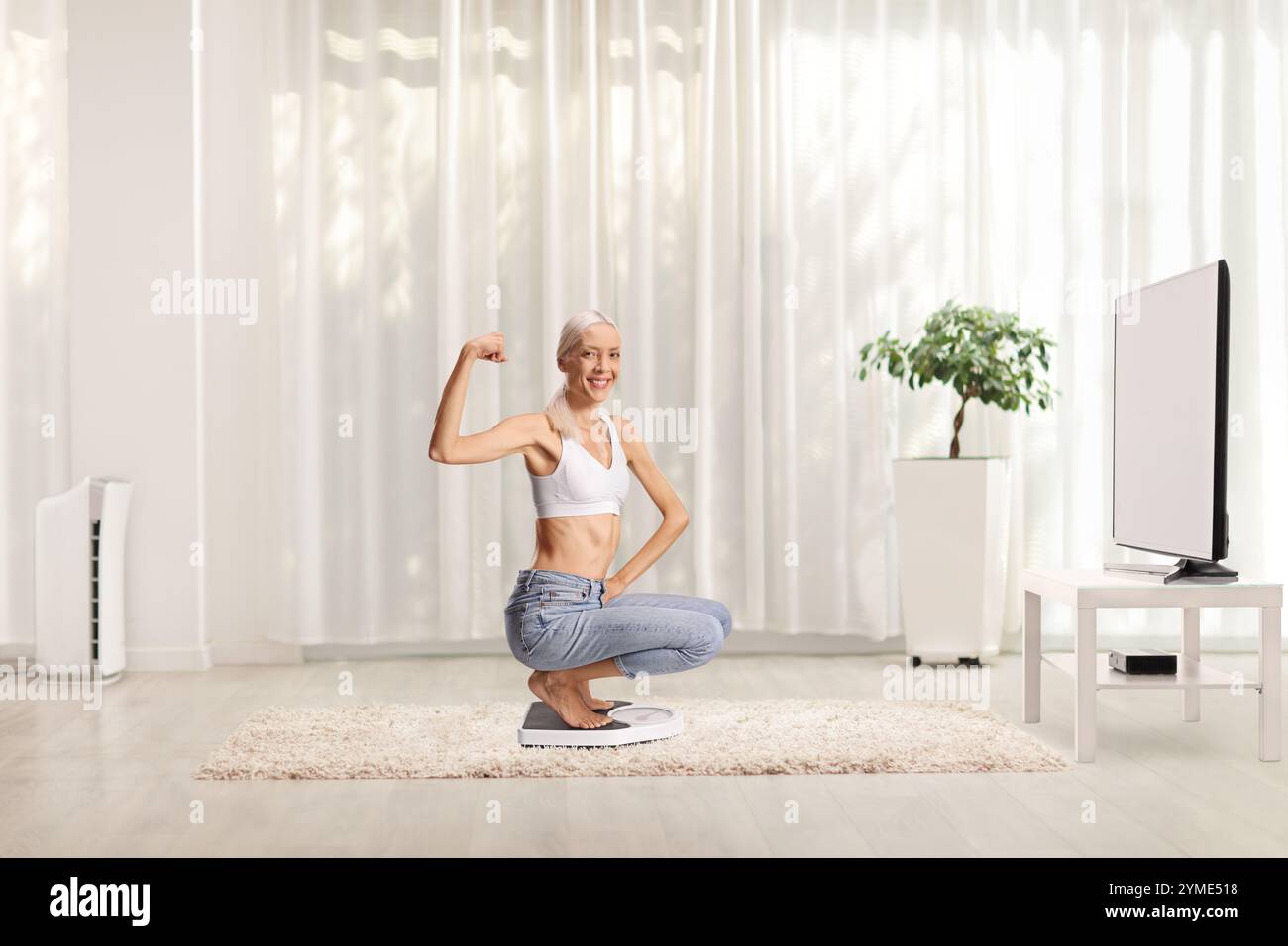 Happy young woman flexing muscle on a weight scale in a room in front ...