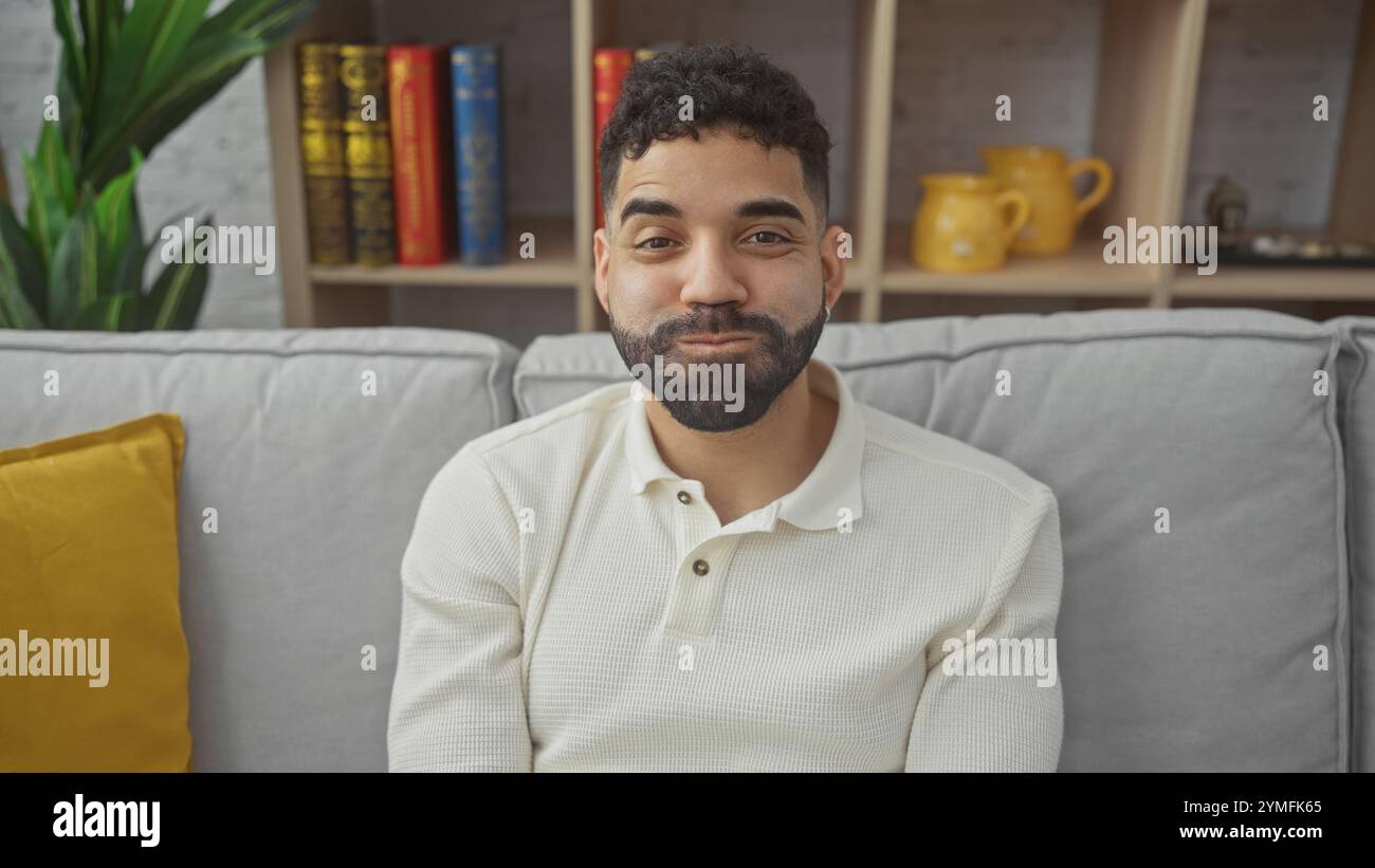 A young hispanic man with a beard smiling casually while seated comfortably on a grey sofa in a ...