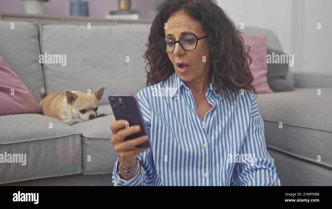 A surprised middle-aged hispanic woman reads a message on her phone beside her resting chihuahua ...