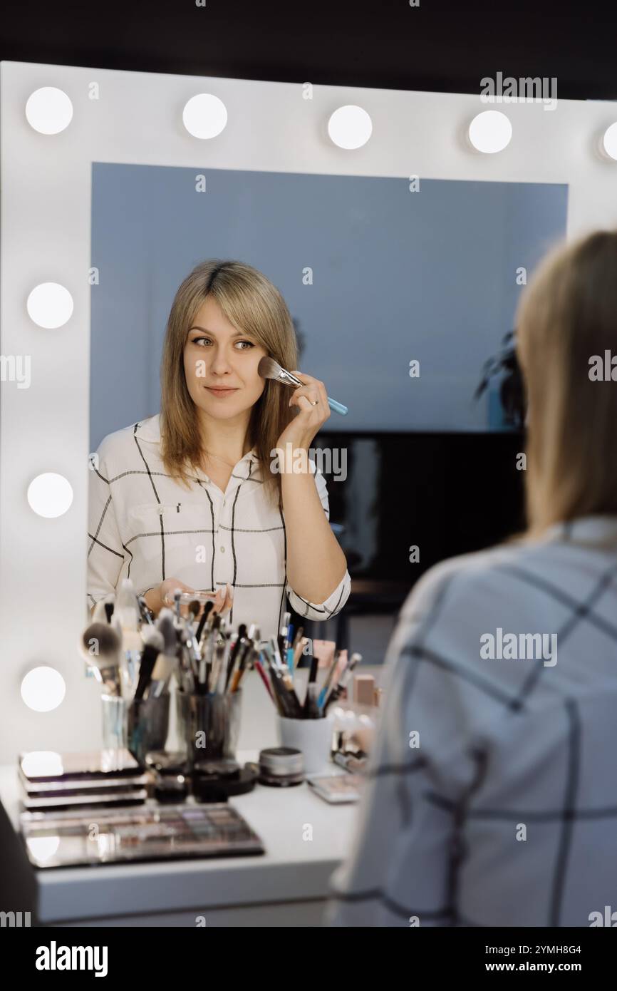 Applying makeup with a brush in front of a lighted mirror for a ...