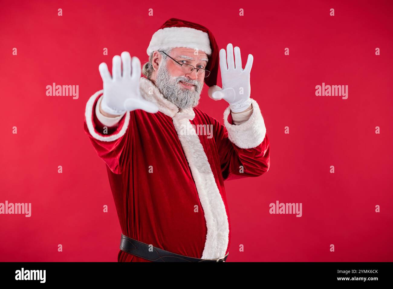 Santa Claus standing against red backdrop gesturing to stop with both ...