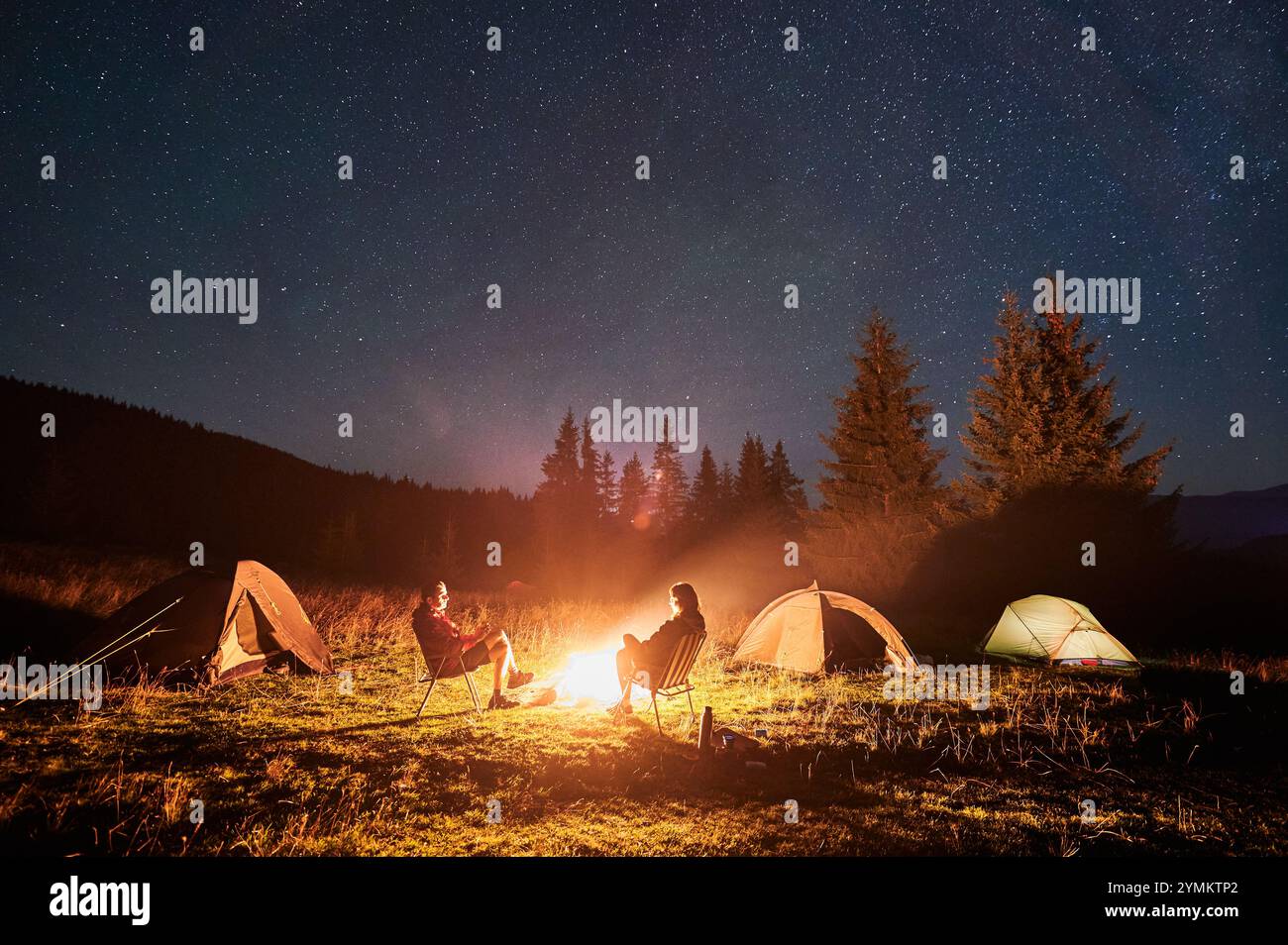 Silhouette of couple hikers resting by campfire under star-studded sky ...