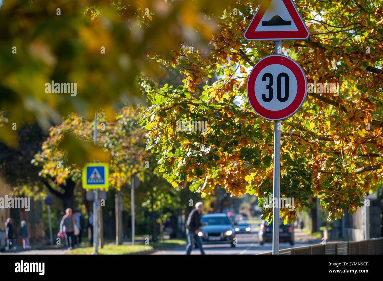 Speed limit signs beside a tree-lined street as pedestrians walk during ...
