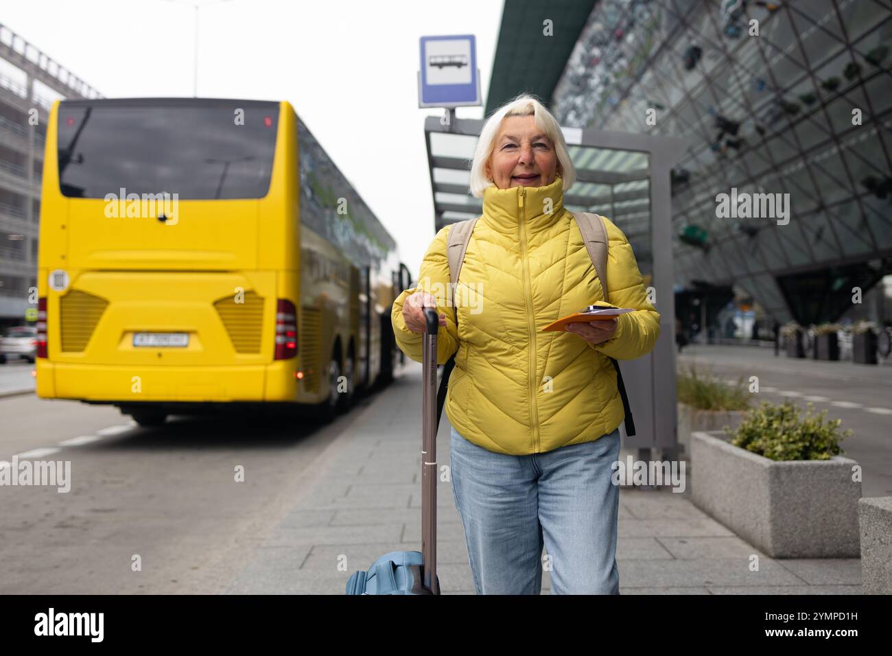 60s senior woman standing on the bus station and using the smartphone ...