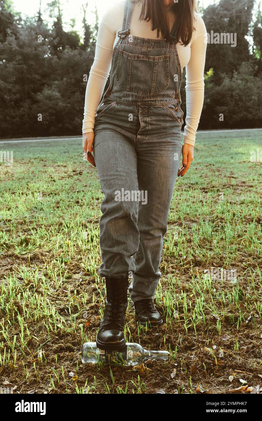 Woman wearing overalls and boots crushing a glass bottle with her foot ...